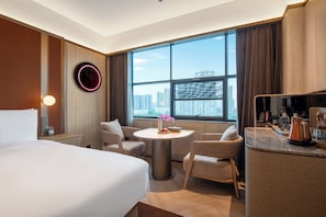 Quarto de hotel com cama, mesa redonda, duas cadeiras, espaço para café e uma grande janela com vista para a cidade.