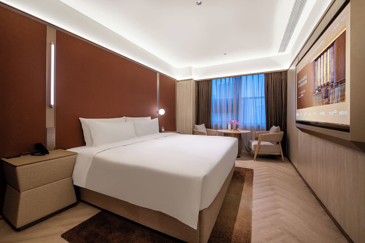Quarto de hotel com uma cama grande, mesa de cabeceira, cadeira e uma pequena mesa com abajur.