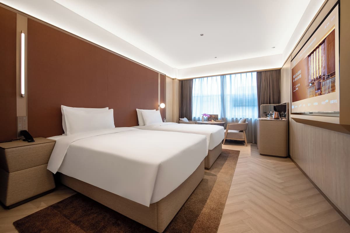 Quarto de hotel com duas camas, uma escrivaninha, uma cadeira, uma televisão e uma janela grande com cortinas.