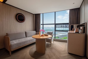 Quarto de hotel moderno com sofá, mesa redonda, cadeira, minibar e uma janela ampla com vista para a cidade.
