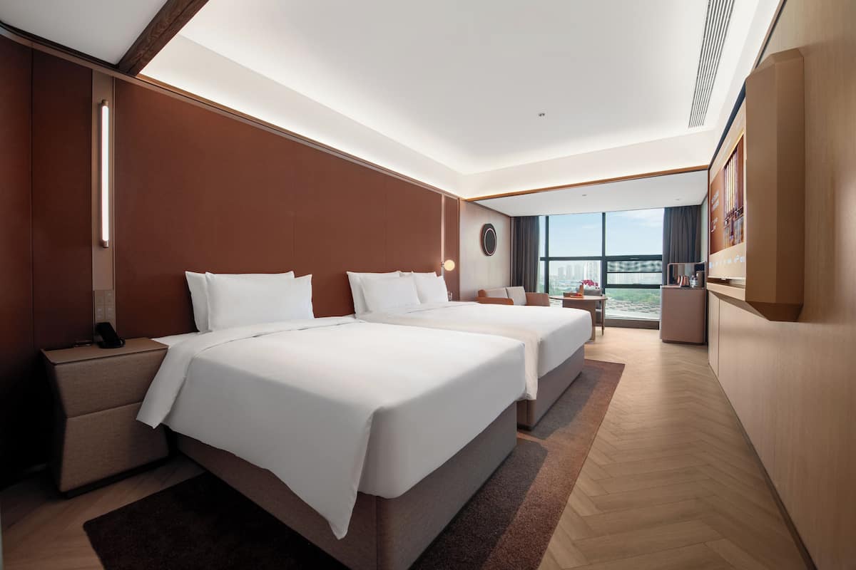 Quarto de hotel com duas camas, uma escrivaninha, uma cadeira, uma mesinha, uma janela com vista e um minibar.