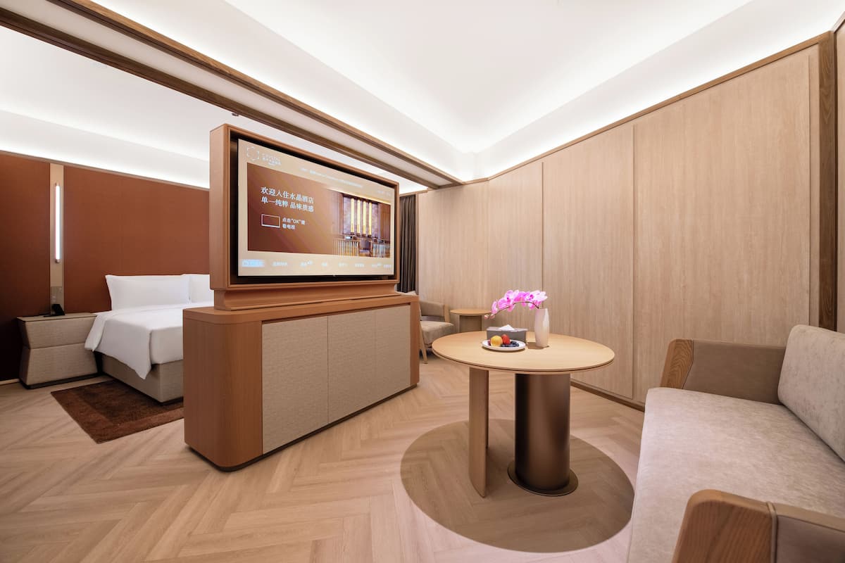Quarto de hotel moderno com uma cama, área de estar, TV fixada na parede e uma pequena mesa com um vaso de flores.