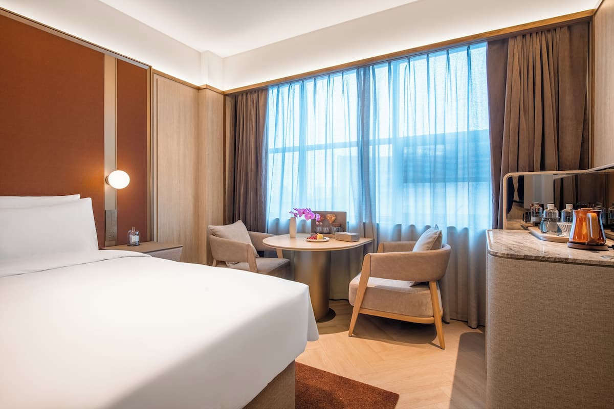 Quarto de hotel com uma cama grande, uma mesa redonda, duas cadeiras, uma pequena escrivaninha, um espelho e uma janela com cortinas.