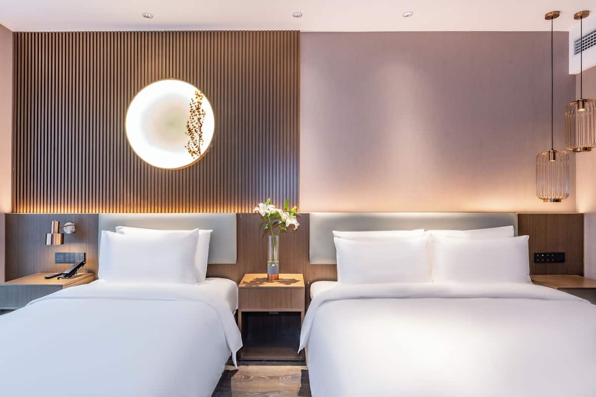 Quarto de hotel com duas camas, uma luminária na parede e uma mesinha de cabeceira com um vaso de flores.