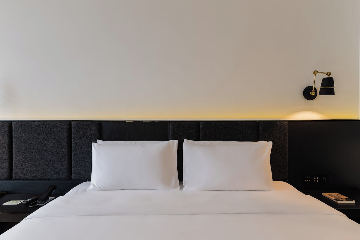 Quarto de hotel com uma cama, uma mesa de cabeceira com abajur e painel de controle fixado na parede.