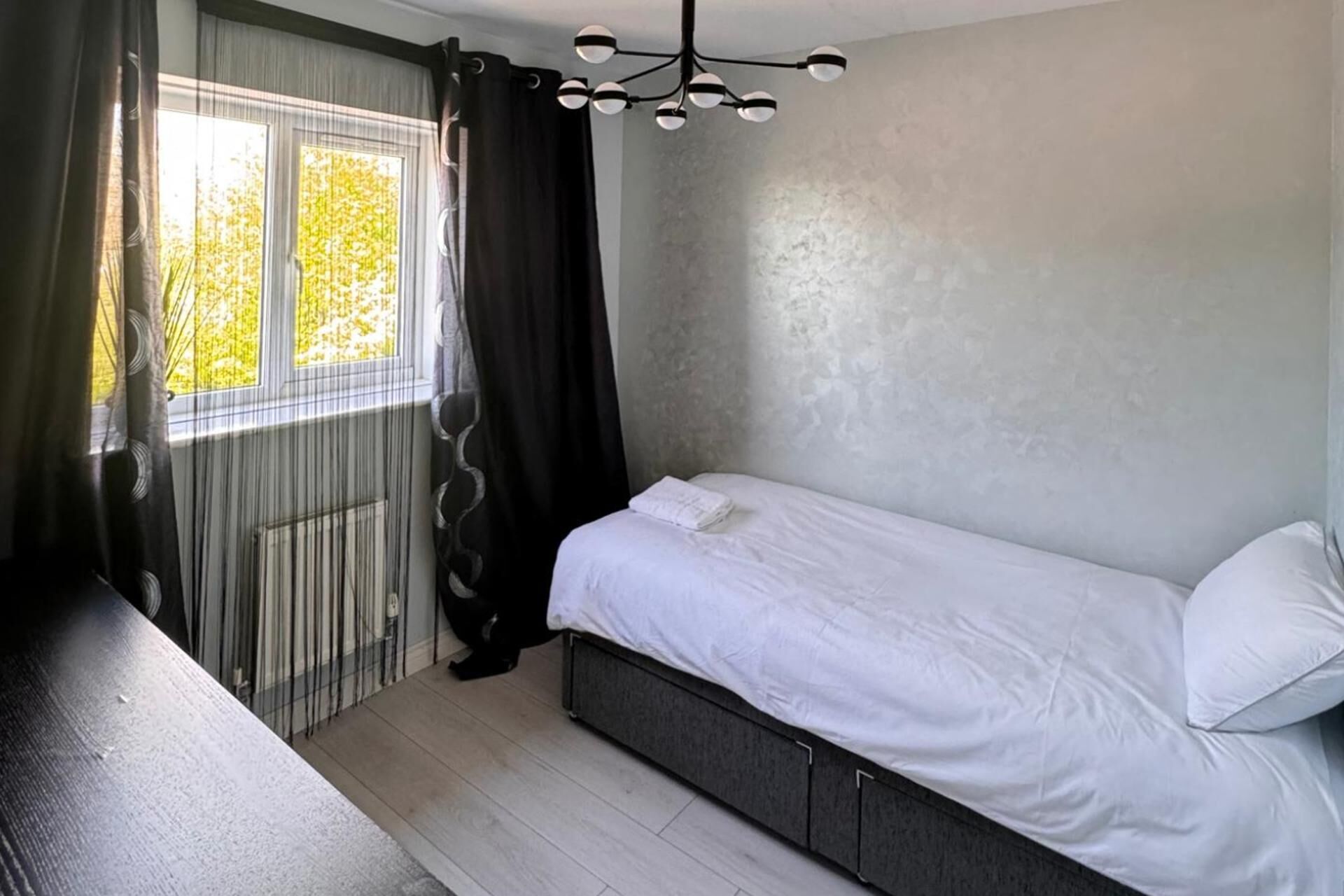 Quarto com uma cama de solteiro, radiador, janela com cortinas e luminária no teto.