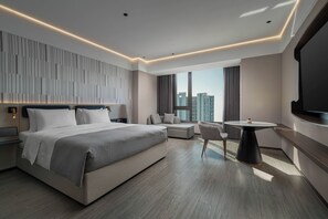 Quarto de hotel moderno com uma cama grande, um sofá pequeno, uma mesa redonda e uma TV fixada na parede.