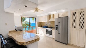 Cozinha com ilha curva, banquetas, geladeira, forno e vista para o oceano através de grandes janelas.