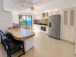 Cozinha com ilha curva, banquetas, geladeira, forno e vista para o oceano através de grandes janelas.