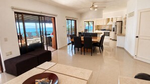 Uma sala de estar moderna com sofá, área de jantar com mesa e cadeiras, cozinha equipada e grandes portas de vidro deslizantes que dão para uma varanda com vista para o oceano.