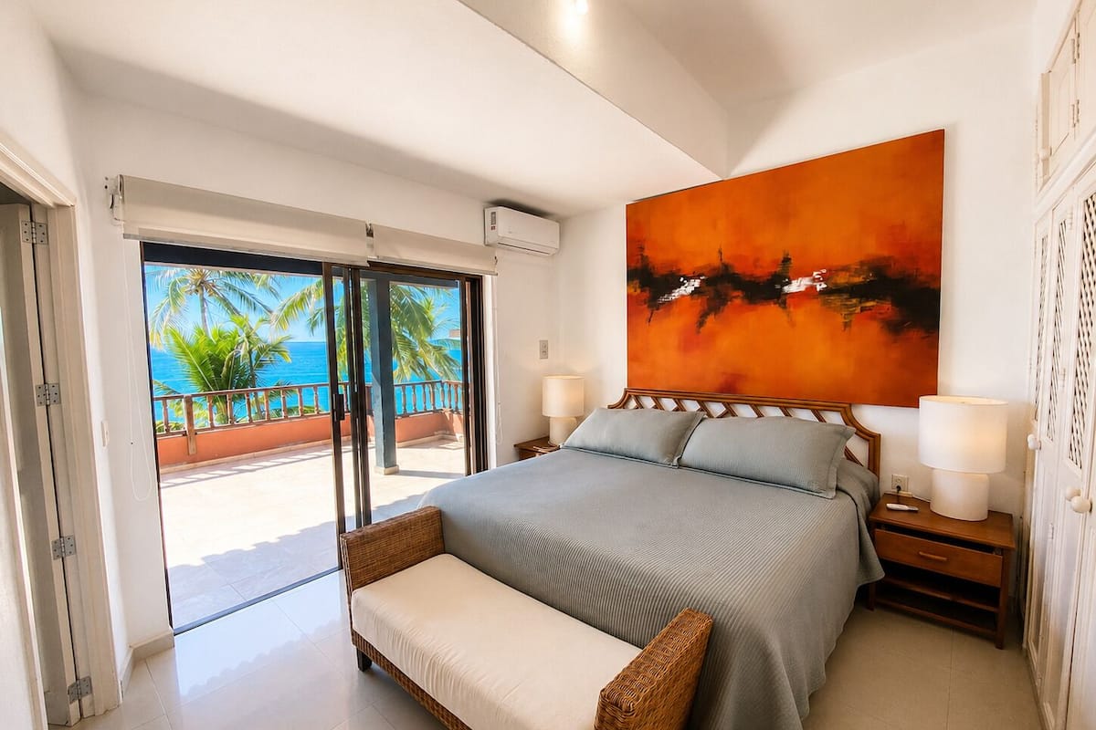 Quarto com cama, um banco, uma pintura e vista para o oceano por uma porta de correr.