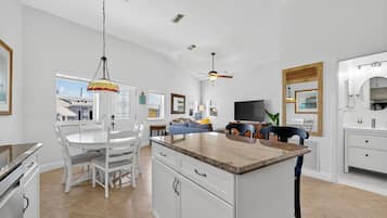 Uma cozinha moderna com armários brancos, uma área de refeições com mesa e cadeiras brancas, um ventilador de teto e uma televisão.