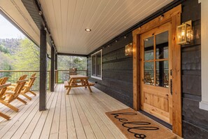 Um deck de madeira com um banco e cadeiras, uma porta de madeira com painéis de vidro e vista para árvores.