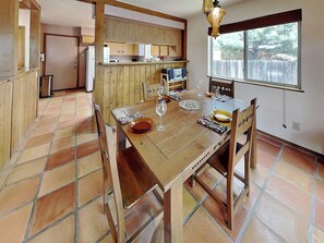 Uma área de refeições com uma mesa de madeira posta para dois, uma cozinha ao fundo e uma janela com vista para árvores.