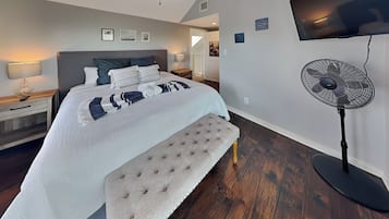 Quarto com cama, mesas de cabeceira, um banco, ventoinha no teto e televisão na parede.