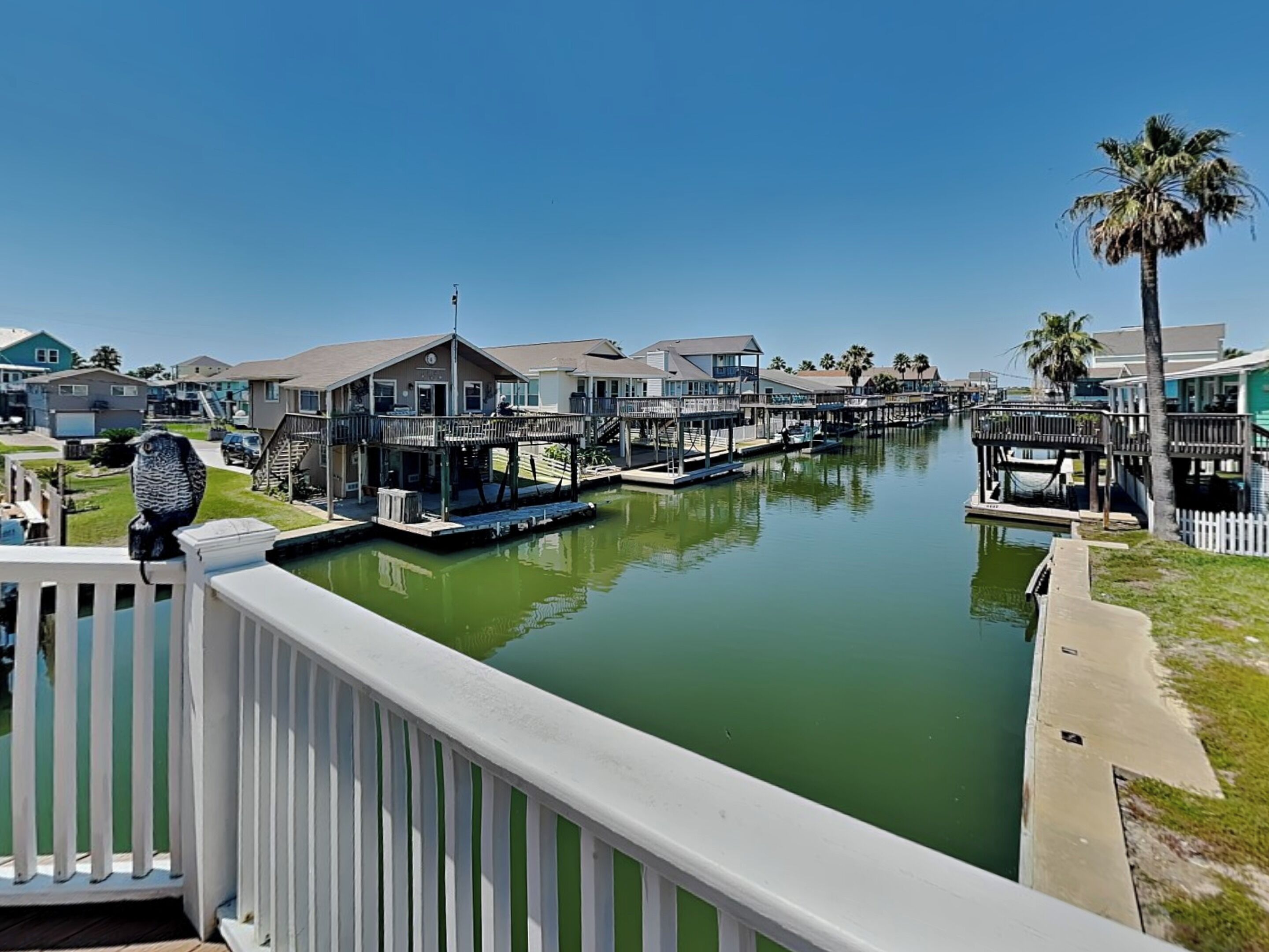 Uma marina com barcos atracados, um corrimão branco e casas ao fundo.