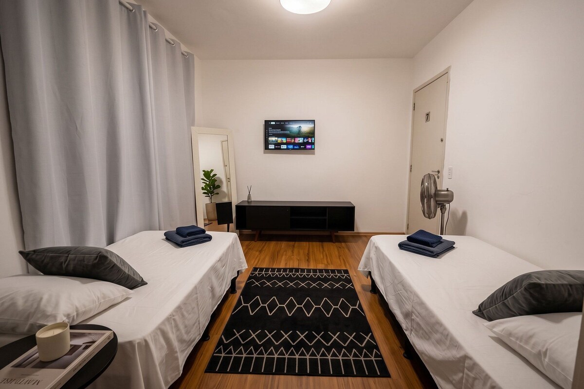 Quarto de hotel com duas camas de solteiro, televisão fixada na parede, mesa de cabeceira com abajur, pequena planta em vaso e tapete com padrão preto e branco.