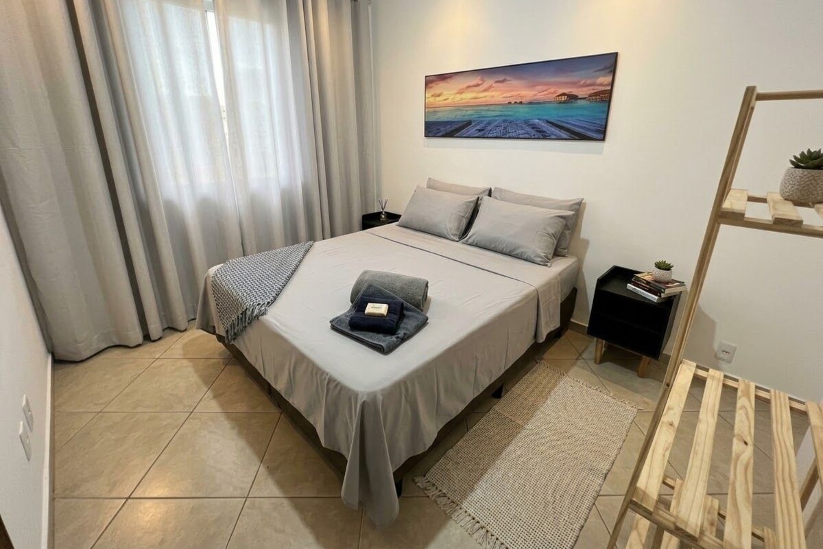 Quarto com cama, mesa de cabeceira, um quadro e uma pequena estante com uma planta.