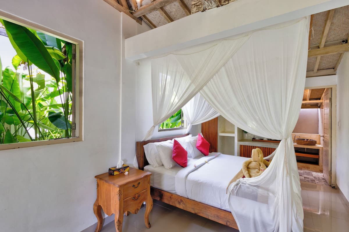 Quarto com cama sob dossel, mesa de cabeceira e janela com vista para vegetação.