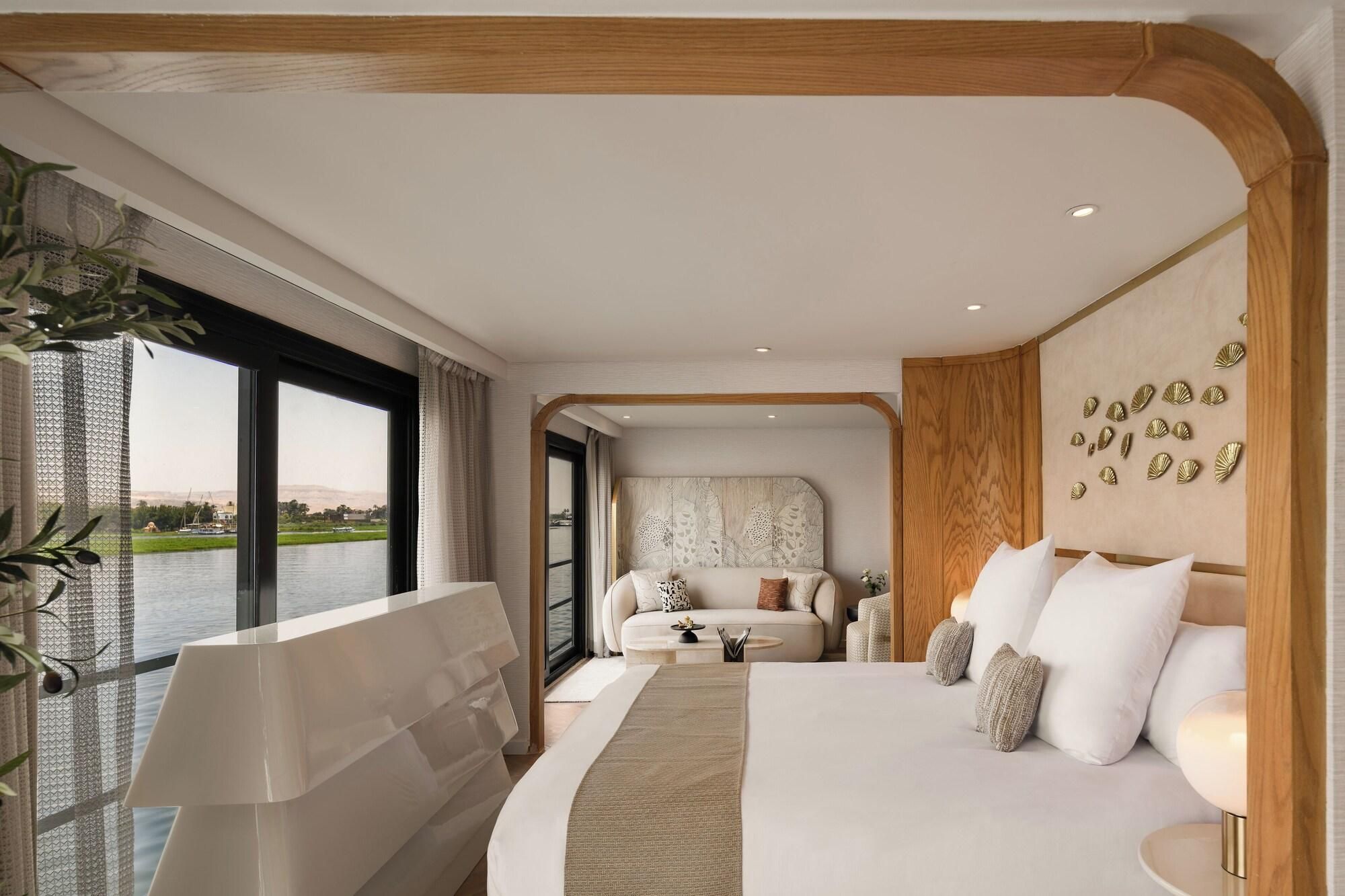 Quarto de hotel moderno com uma cama grande, um sofá, vista para um corpo de água e uma parede decorativa.