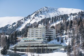 Um hotel de vários andares com design moderno, localizado em uma área montanhosa coberta de neve e com céu claro.
