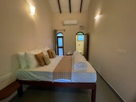 Quarto de hotel com cama, almofadas, um banco, toalha, janela e unidade de ar condicionado.