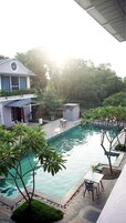 Piscina cercada por vegetação, um terraço com cadeiras e um edifício com janela peculiar.