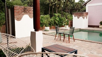 Uma área junto à piscina com mesa e cadeiras, uma cadeira branca, um banco de madeira e uma parede de tijolos.
