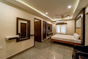 Quarto de hotel com cama, cadeira, escrivaninha, espelho, ventoinha no teto e ar condicionado.