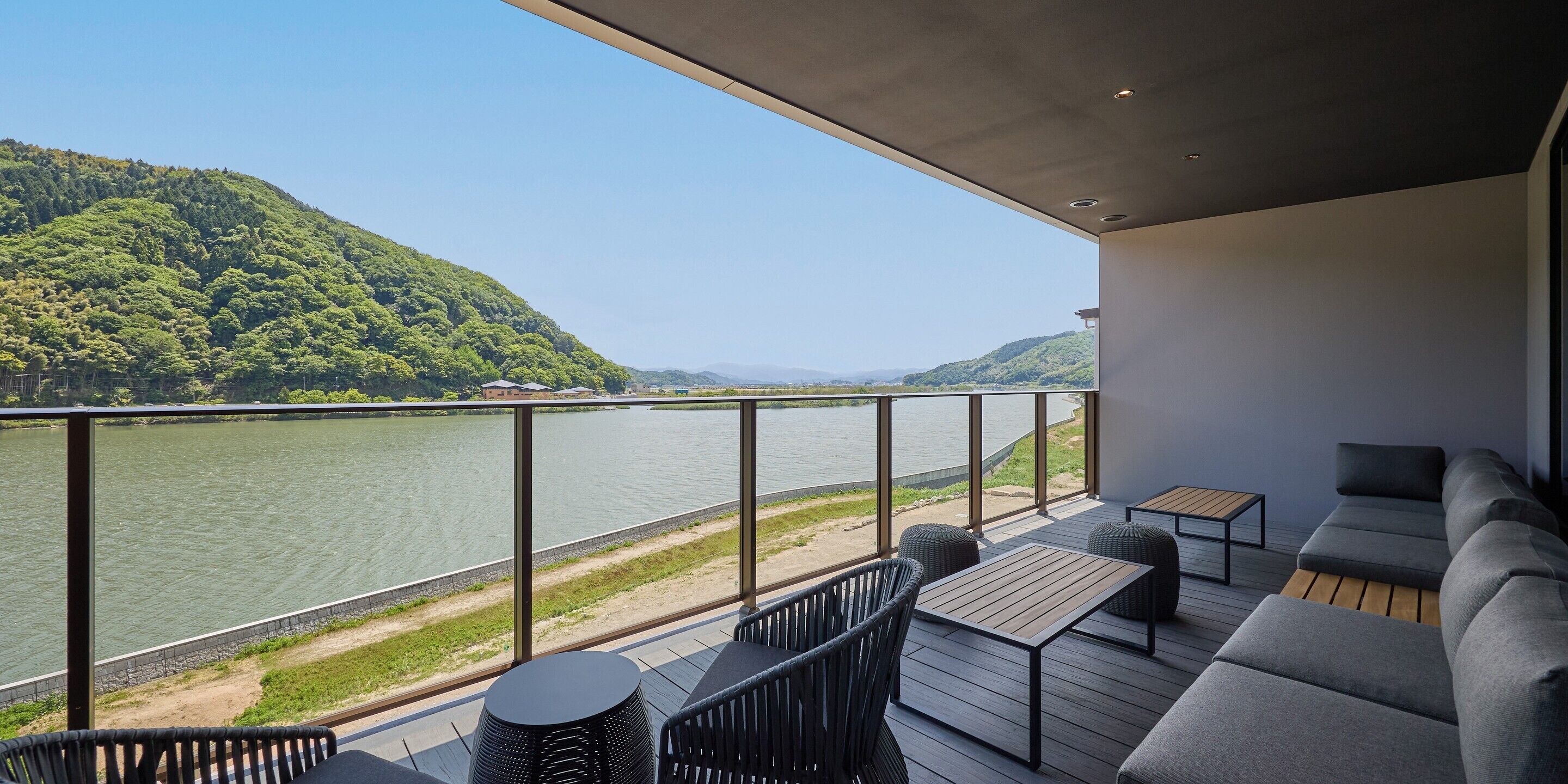 Uma varanda com vista para um rio e colinas, mobiliada com um sofá, cadeiras e uma mesa.