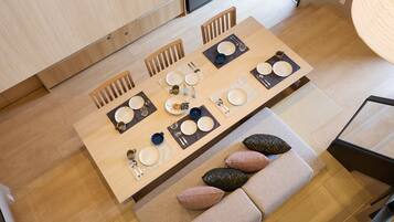 Uma sala de jantar com uma mesa de madeira posta para quatro pessoas, rodeada por cadeiras e um sofá com almofadas.