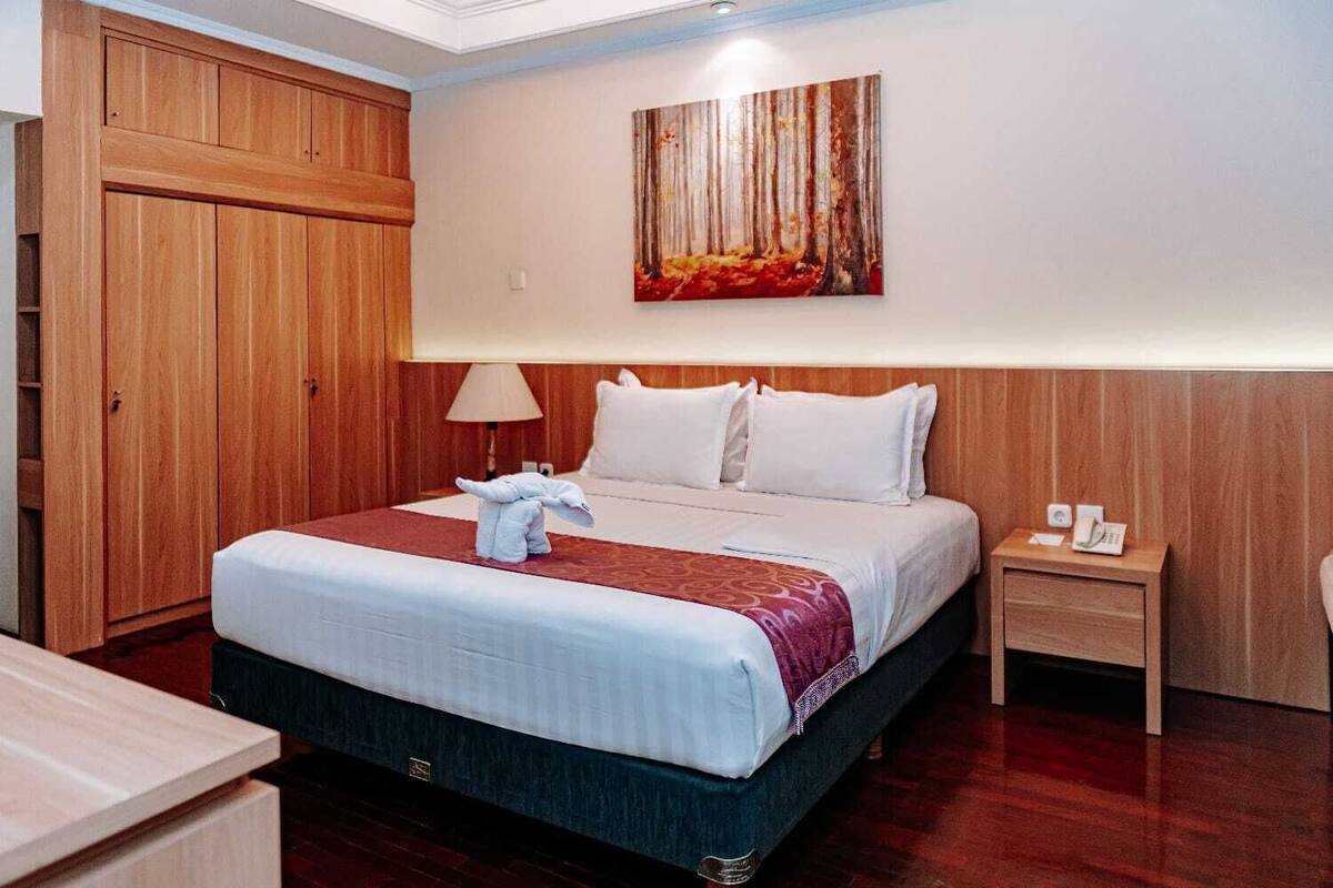 Quarto de hotel com cama grande, guarda-roupa de madeira, mesa de cabeceira, abajur e um quadro na parede.
