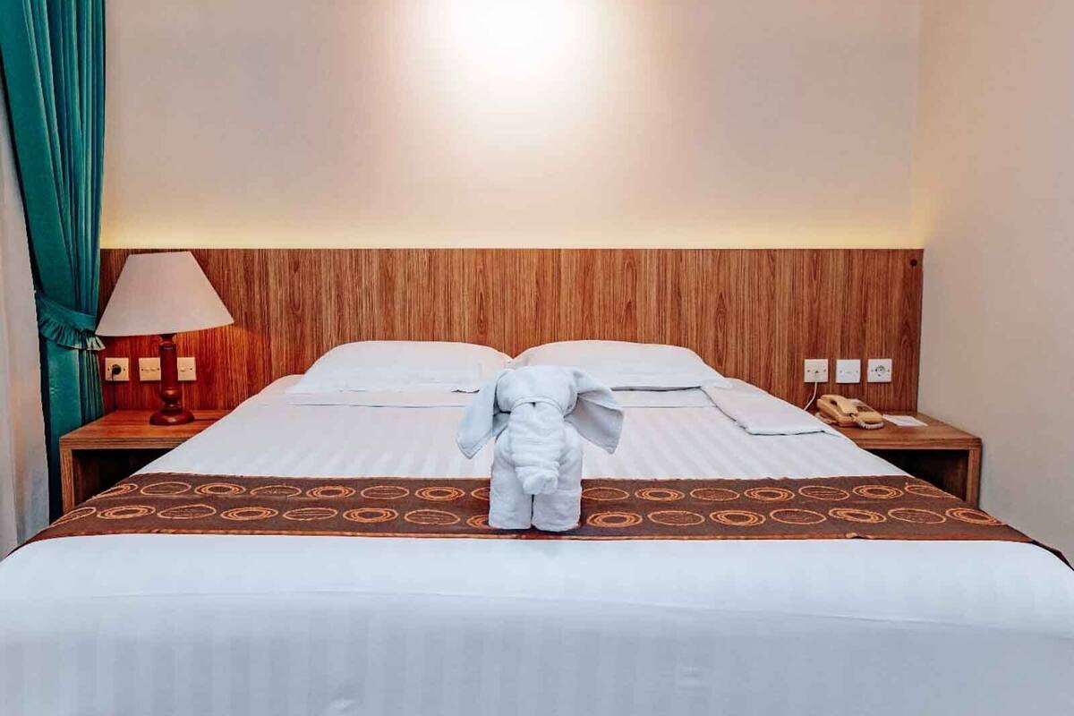 Quarto de hotel com cabeceira de madeira, cama com colcha estampada, abajur, telefone e um brinquedo macio sobre a cama.
