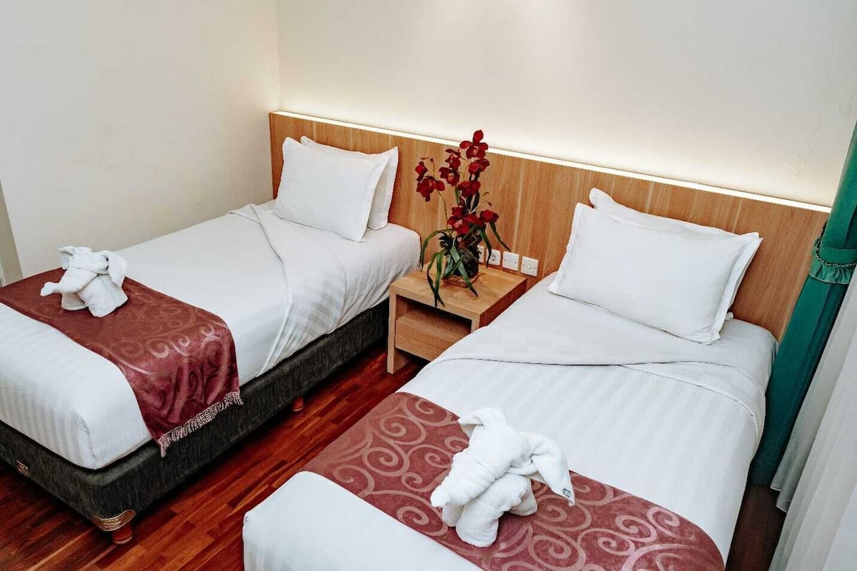 Quarto de hotel com duas camas, cada uma com lençóis brancos e uma colcha vermelha estampada. Há uma mesa de cabeceira de madeira entre as camas, com um vaso de flores vermelhas.