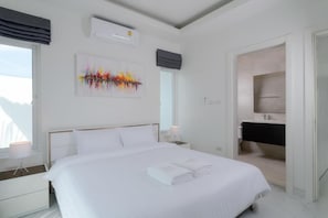 Quarto de hotel com cama, mesa de cabeceira, ar condicionado, banheiro com espelho e pia, e uma pintura abstrata colorida na parede.