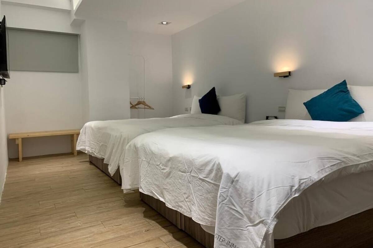 Quarto de hotel com duas camas, um banco, uma televisão e um candeeiro fixado na parede.