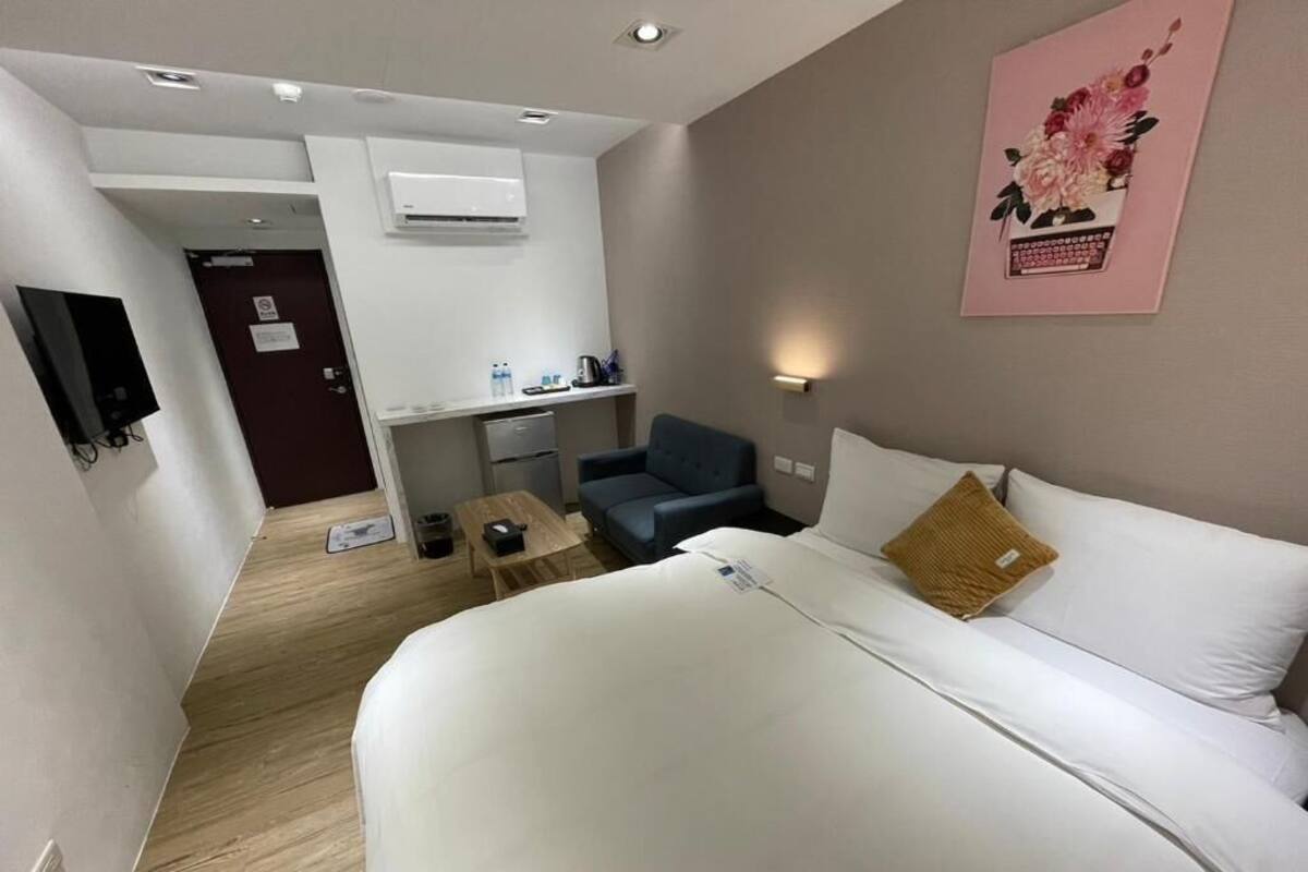 Quarto de hotel com cama, escrivaninha, cadeira, mesa pequena e televisão na parede.