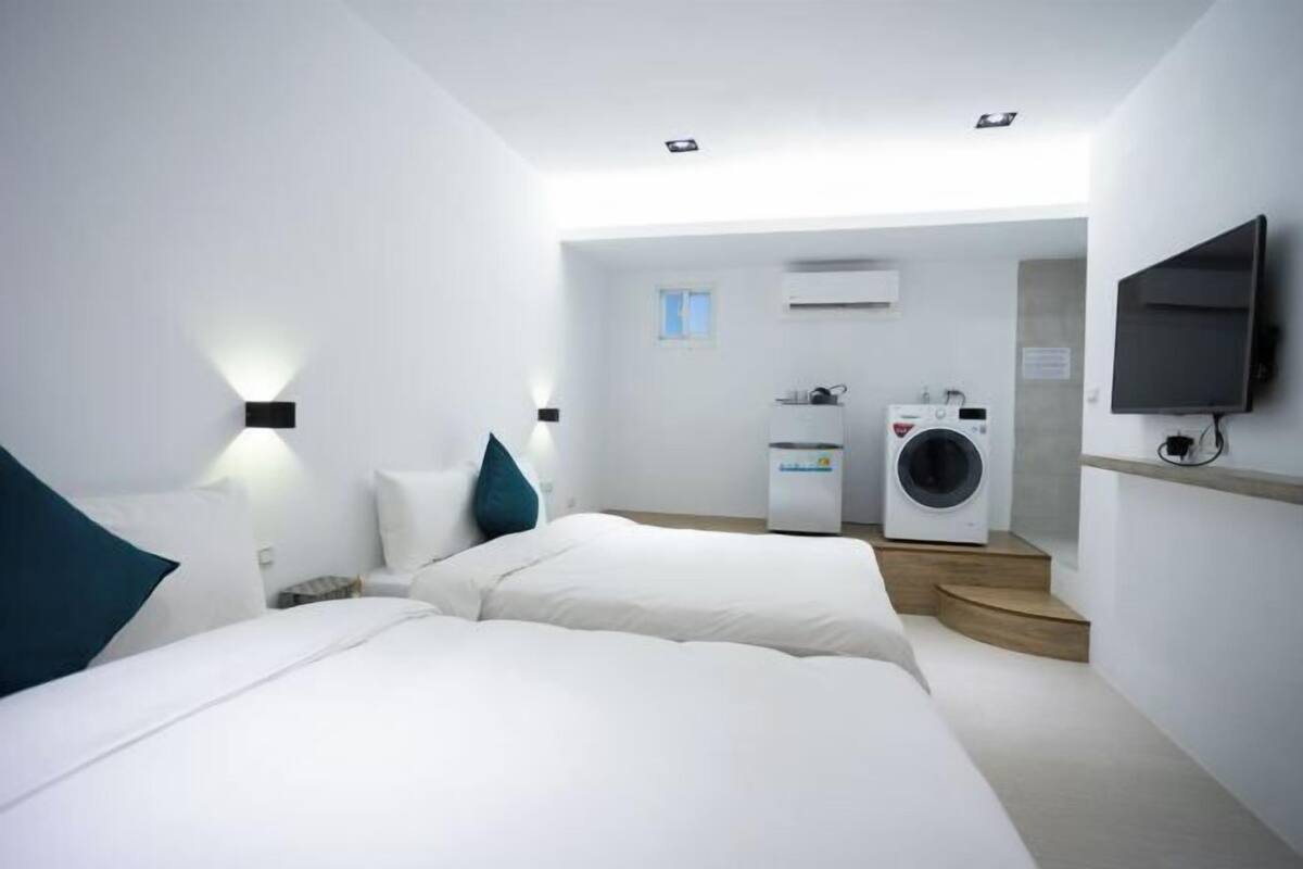 Quarto de hotel moderno com duas camas, máquina de lavar, televisão e uma pequena cozinha.