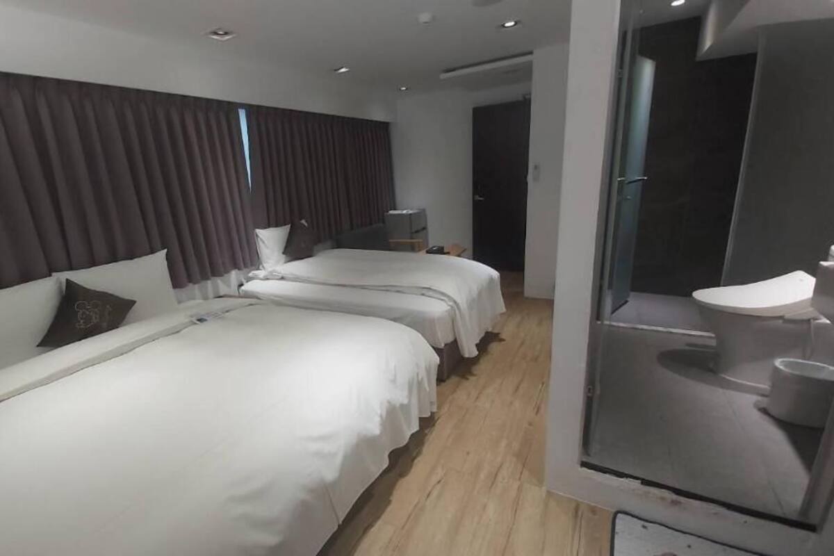 Quarto de hotel com duas camas, uma escrivaninha, uma cadeira e banheiro com vaso sanitário e pia.