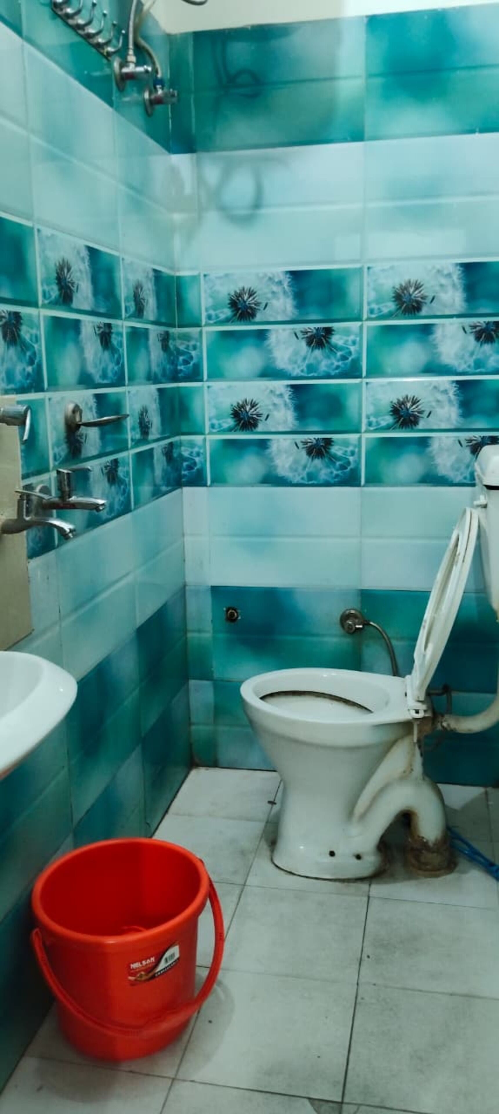 Um banheiro com azulejos azuis, um vaso sanitário branco, um balde vermelho e uma pia.