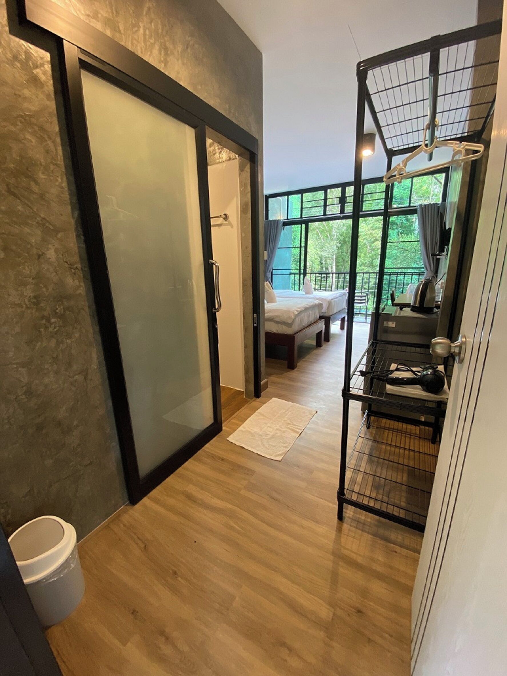 Corredor com porta deslizante que dá para uma varanda com cama e mesa. Há um suporte de metal com uma xícara.
