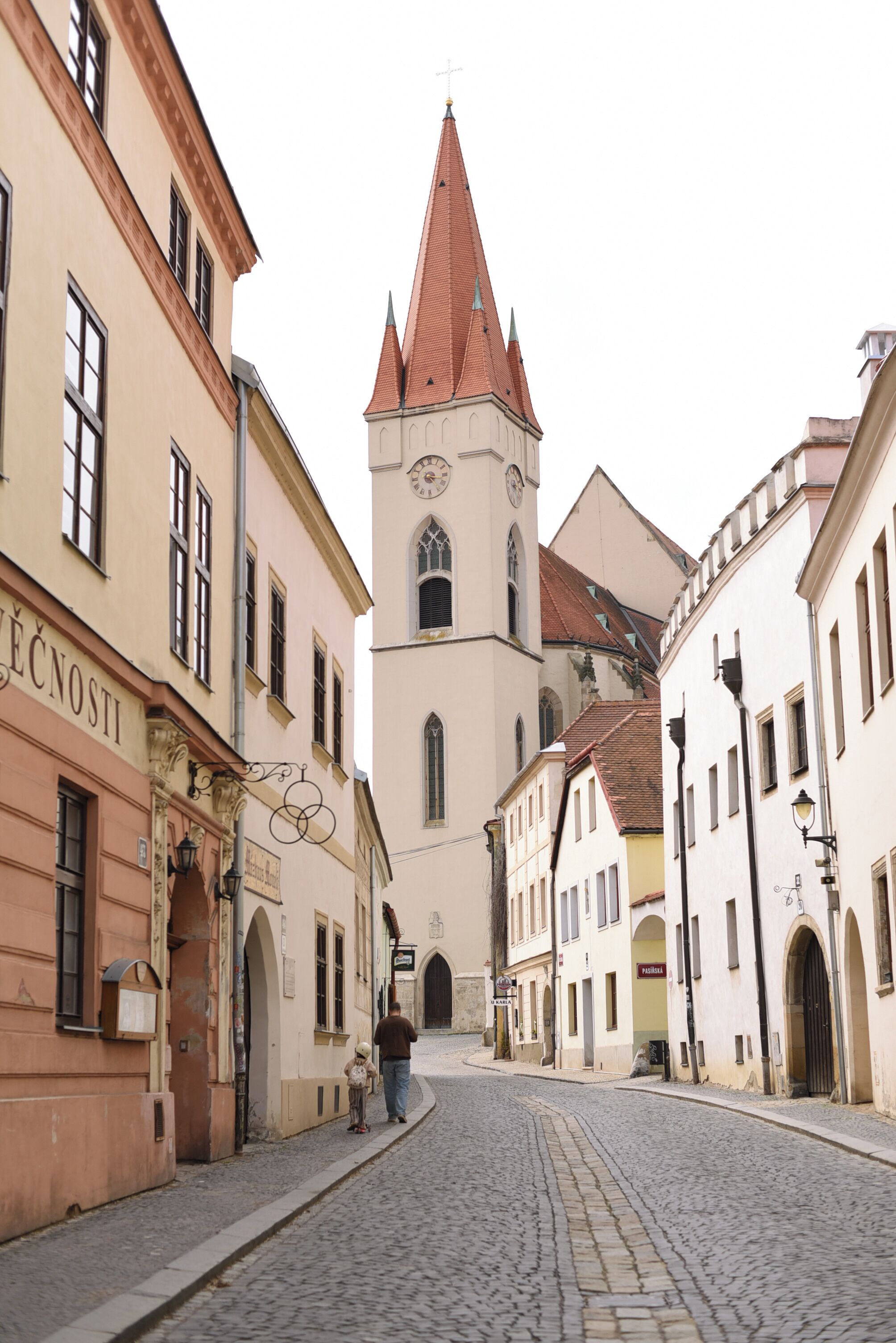 Uma rua de paralelepípedos flanqueada por edifícios históricos, uma igreja com telhado vermelho e uma placa com a inscrição 'WEČNOSTI'.