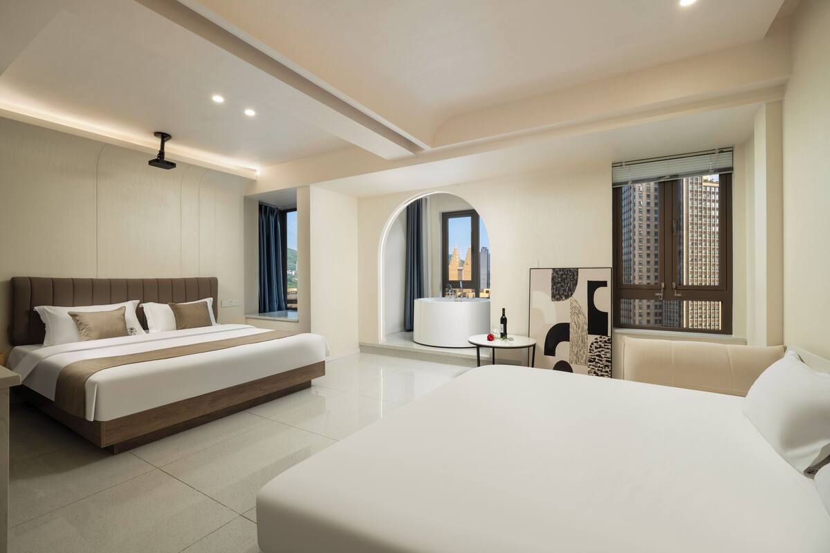 Quarto de hotel moderno com uma cama grande, uma mesa redonda pequena, um sofá e vista para a paisagem urbana.