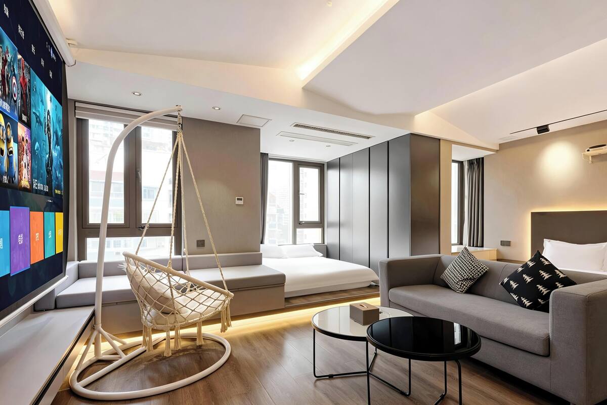 Quarto de hotel moderno com sofá, cadeira suspensa, cama, televisão e uma janela grande.