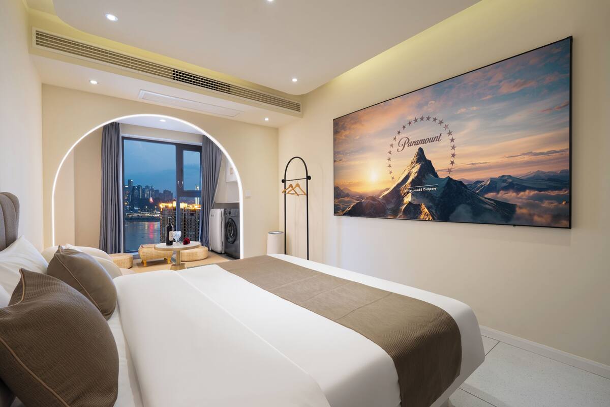 Quarto de hotel com uma cama grande, vista para a cidade, um pôster de filme Paramount e uma luminária circular.