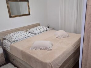 Cama bem arrumada com colcha bege e duas almofadas, cabeceira de madeira, mesa de cabeceira com abajur e espelho na parede.