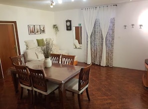 Uma área de jantar com uma mesa e cadeiras de madeira, um vaso com flores, um relógio na parede e uma janela com cortinas.