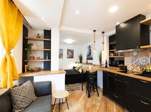 Uma cozinha compacta com área de refeições, armários pretos, bancadas de madeira e um pequeno espaço de estar com sofá e mesa redonda.