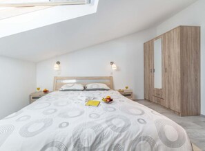 Quarto com teto inclinado, cama com colcha estampada, mesas de cabeceira com abajures e um guarda-roupa de madeira.