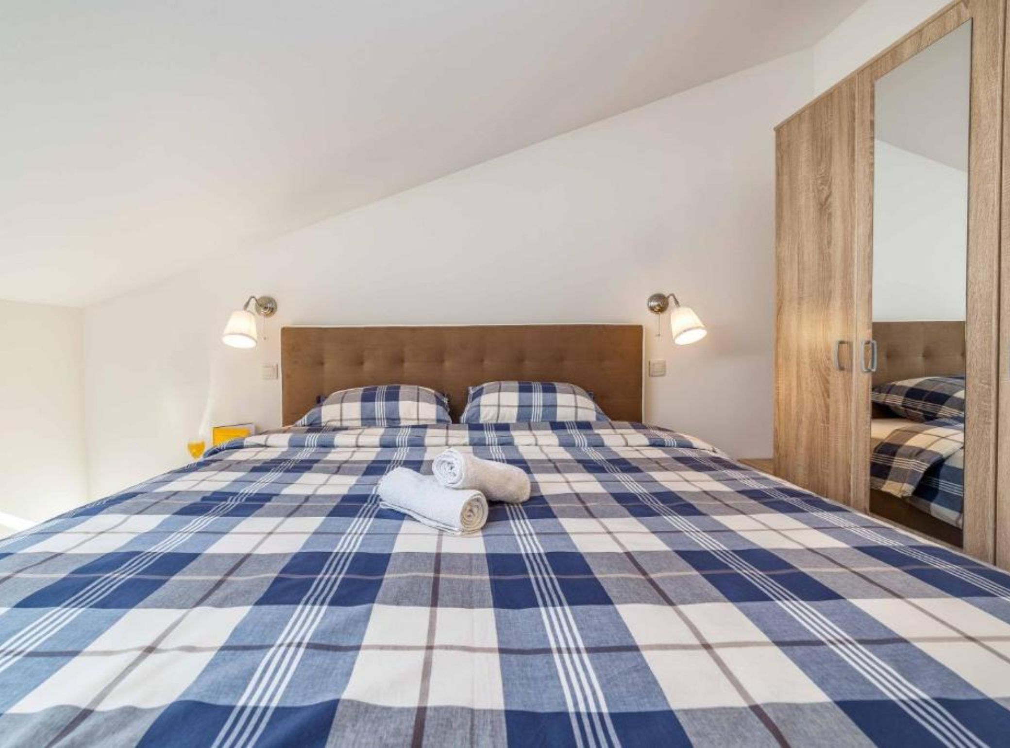 Quarto com colcha xadrez, cabeceira de madeira, duas luminárias de apoio e um guarda-roupa com espelho.